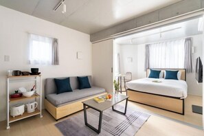 Quarto de hotel moderno com sofá, cama, mesa pequena e área de cozinha compacta.