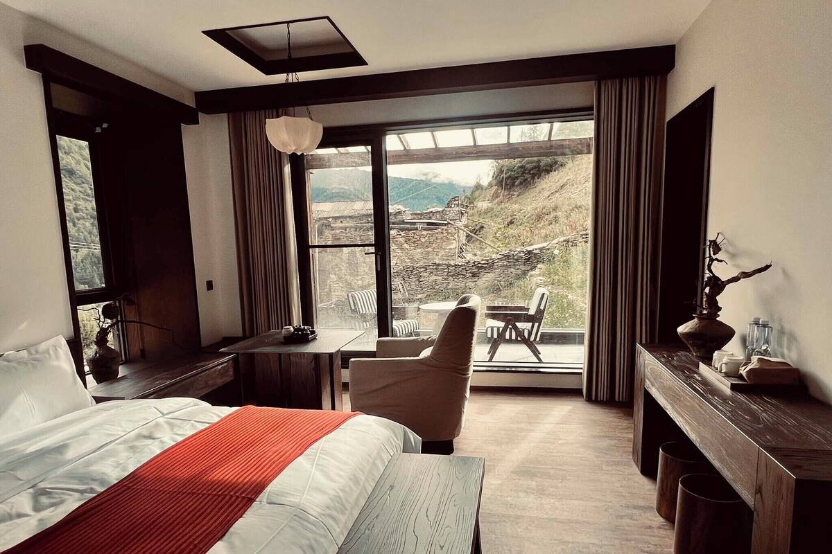 Quarto de hotel com uma cama grande, uma escrivaninha, uma cadeira e vista para uma paisagem montanhosa pelas janelas.