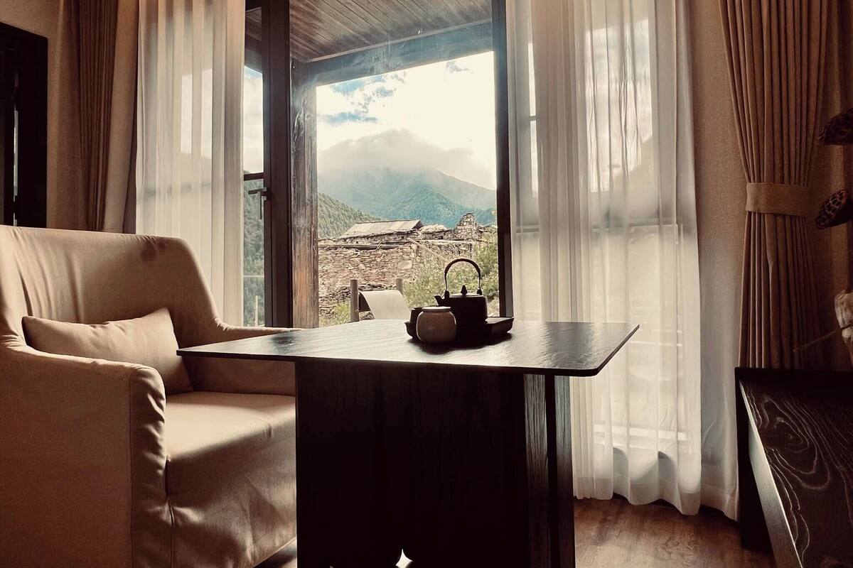 Quarto com vista para montanhas através de uma janela ampla, uma mesa com um bule e um sofá.