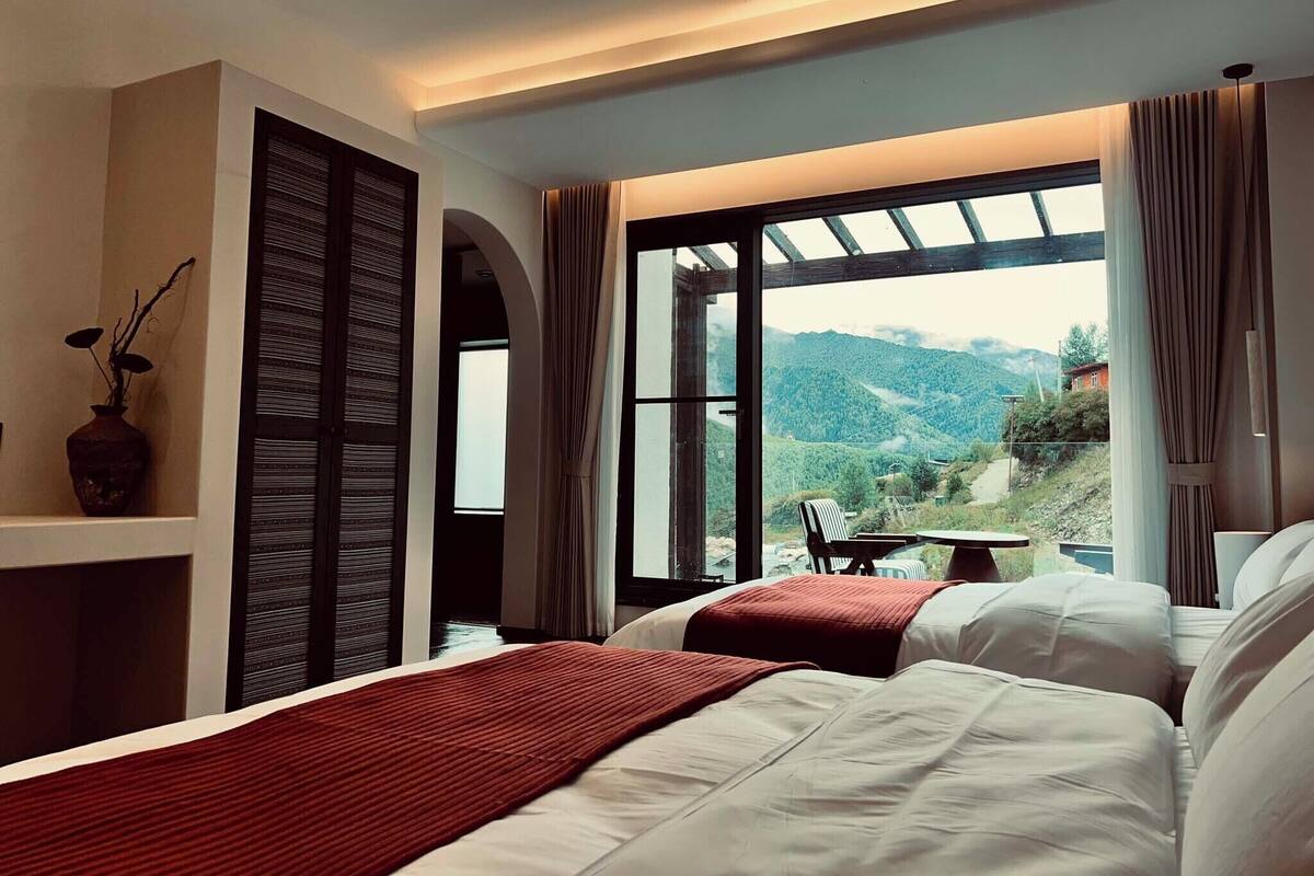 Quarto de hotel com duas camas, uma janela grande com vista para as montanhas e uma pequena mesa com uma cadeira no exterior.