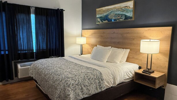 Quarto de hotel com uma cama, duas luminárias de cabeceira, uma cabeceira de madeira, um quadro na parede e uma janela com cortinas.