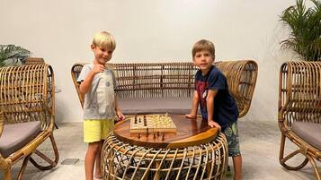 Duas crianças jogando xadrez em uma mesa redonda, cercada por cadeiras de vime.