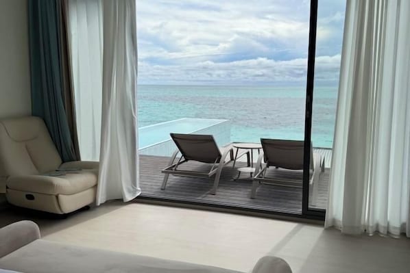 Quarto com uma grande janela com vista para uma praia. Há um sofá e uma pequena mesa com duas cadeiras.