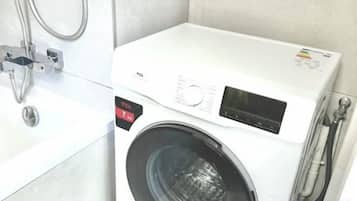 Máquina de lavar com carga frontal, painel de controle, mangueira de entrada de água e tubulação metálica.