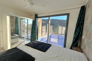 Quarto com cama, ventilador de teto, porta de vidro deslizante para uma varanda e vista para uma piscina e edifícios.