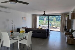 Uma sala de estar moderna com uma mesa de jantar de vidro, cadeiras brancas, um sofá escuro e uma grande porta de vidro deslizante que dá para uma área externa.