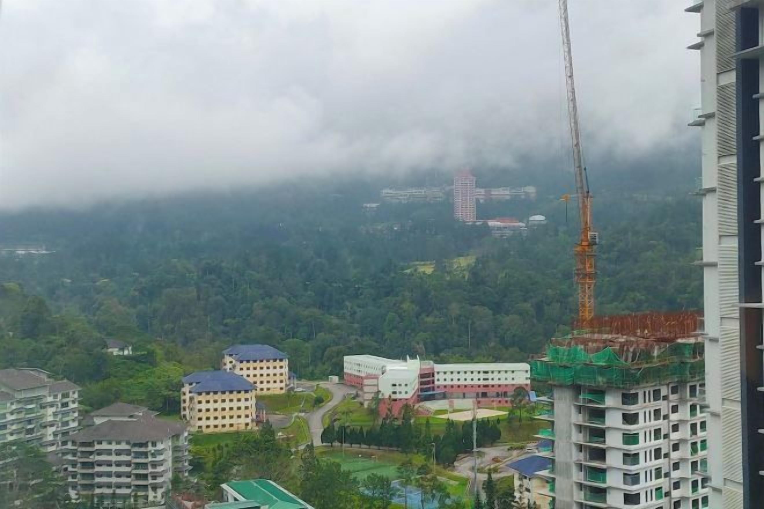 Vista de um canteiro de obras com edifícios e uma grua, cercado por vegetação e céu nublado.
