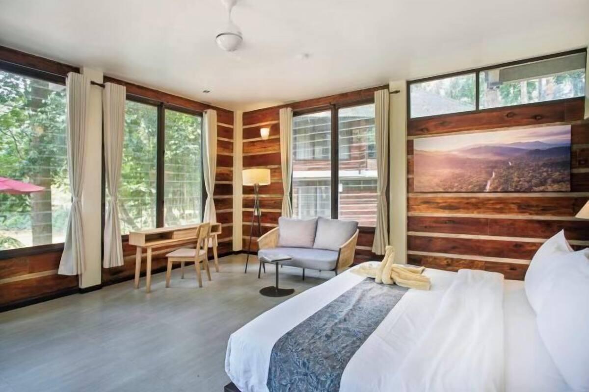 Um quarto com cama, uma cadeira, uma escrivaninha e janelas amplas.