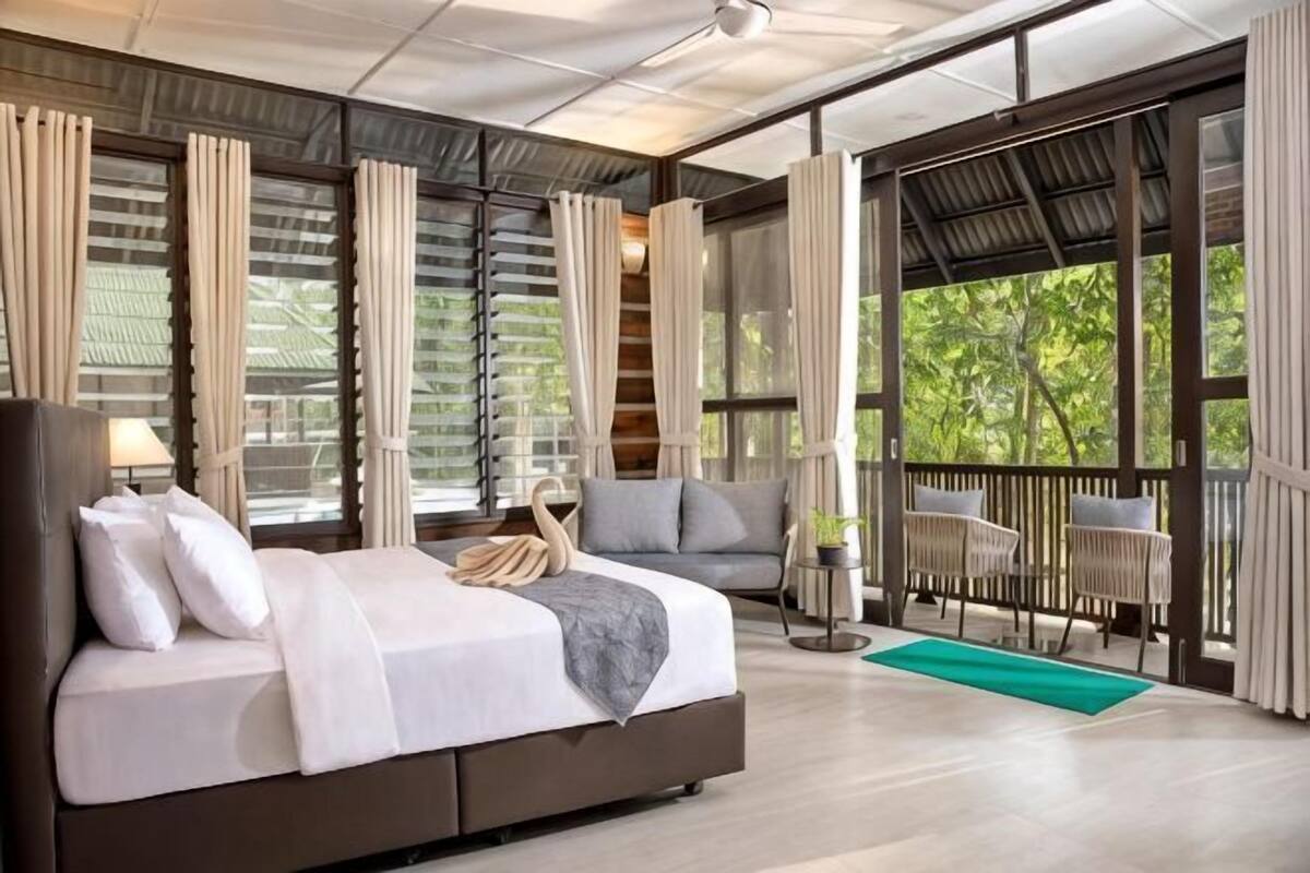 Quarto moderno com cama grande, um sofá, uma mesinha e vista para o verde através de amplas janelas.