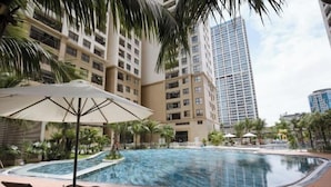 Uma piscina com espreguiçadeiras e guarda-sóis, cercada por palmeiras e edifícios altos.