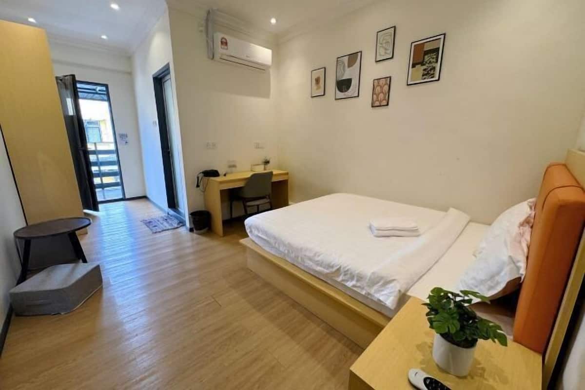 Quarto de hotel moderno com piso de madeira, cama com roupa branca, escrivaninha com cadeira, pequena mesa com planta e ar-condicionado fixado na parede.