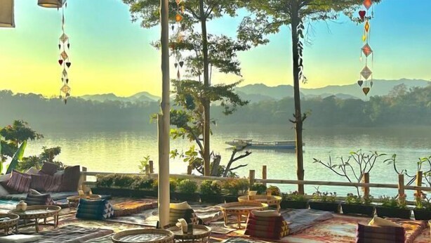 Uma área de estar ao ar livre tranquila, com vista para um lago e montanhas, equipada com almofadas e mesas.
