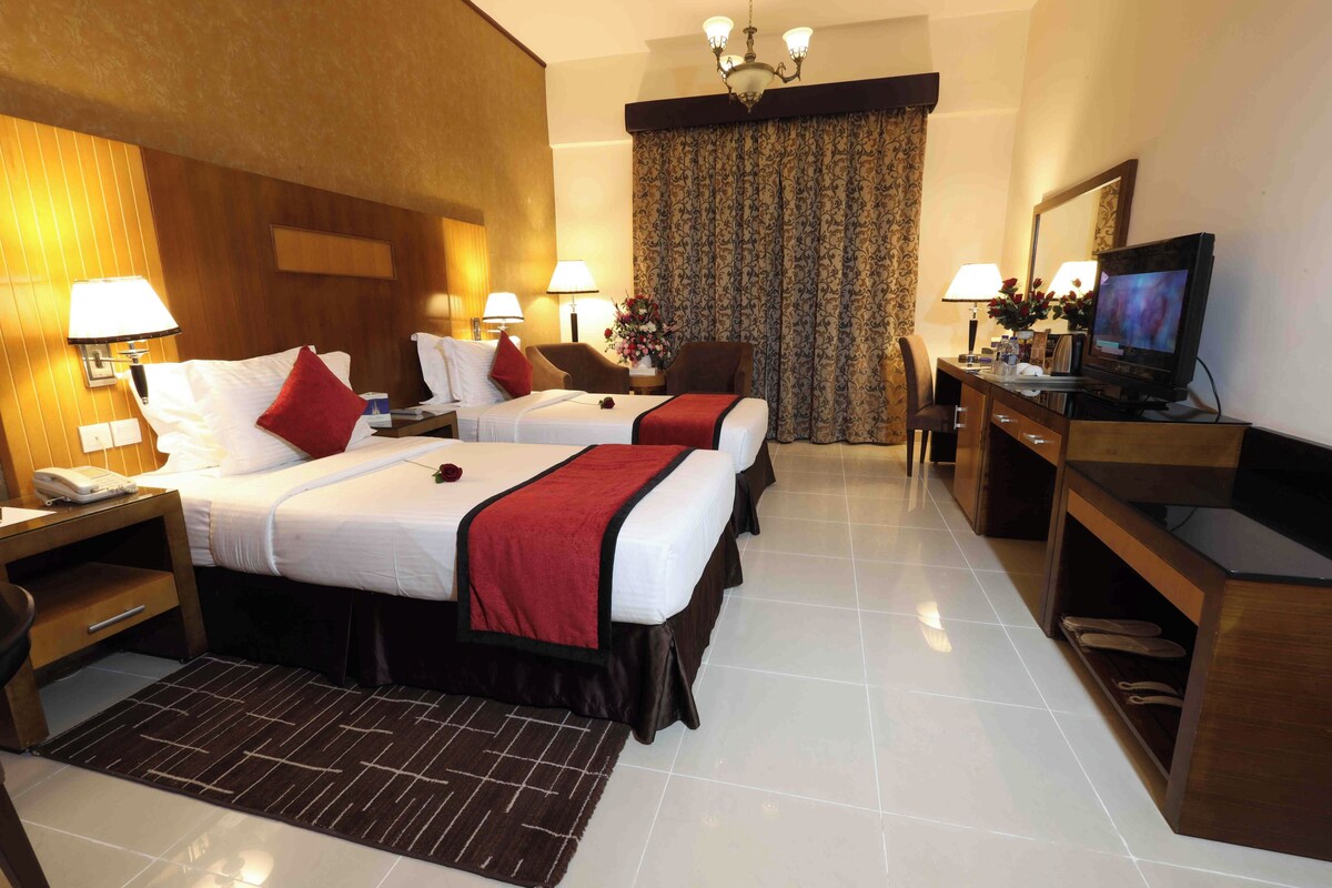 Quarto de hotel com duas camas, uma escrivaninha com televisão e banheiro.