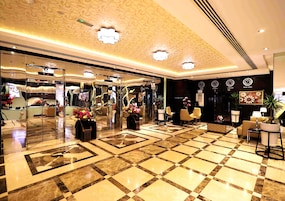 Um saguão de hotel com piso xadrez, um balcão de recepção e uma parede com relógios mostrando diferentes cidades.