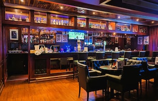 Uma área de bar com prateleiras de madeira repletas de garrafas, uma tela de televisão e assentos de couro.