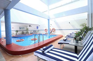 Piscina interna com espreguiçadeiras e um solário, cercada por um espaço revestido e teto de vidro.