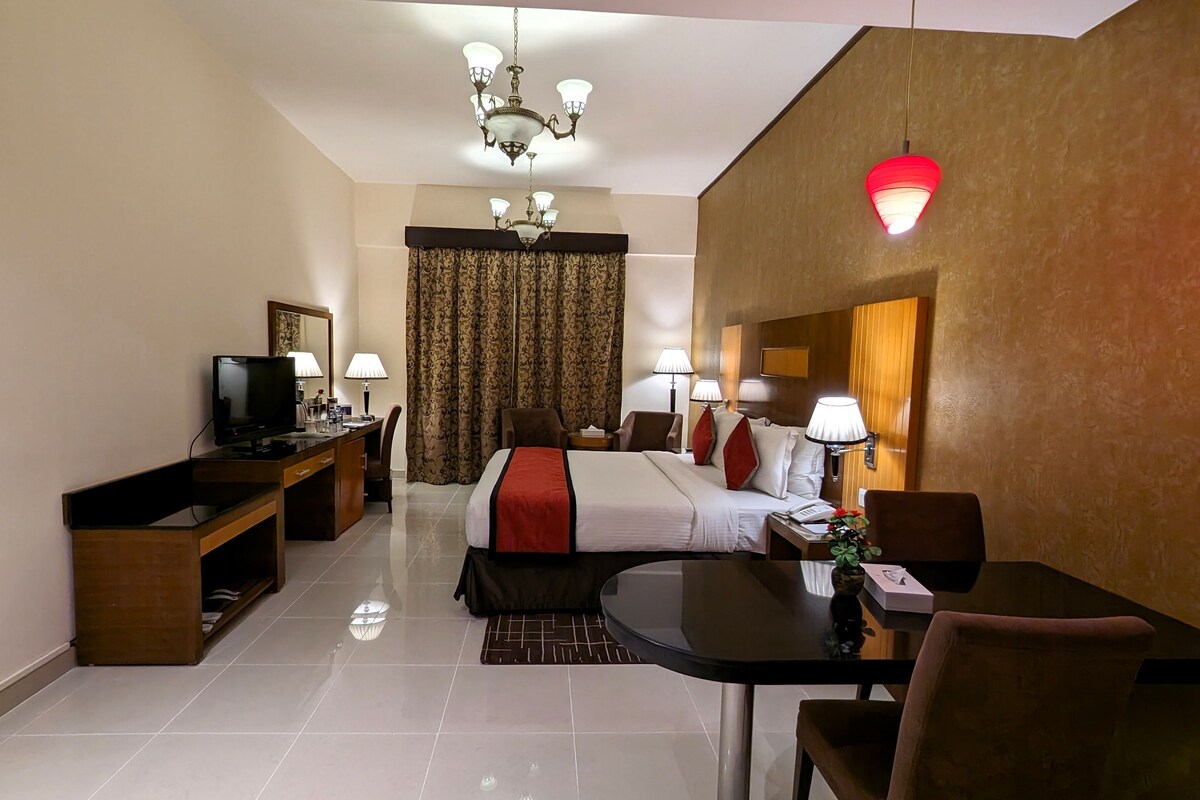 Quarto de hotel com cama, escrivaninha, televisão, luminárias e um candelabro.