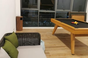 Quarto com mesa de bilhar, sofá de vime com almofadas e janela com vista para a cidade à noite.