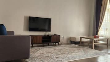 Uma sala de estar com televisão de tela plana na parede, um sofá, uma mesa de centro e um tapete estampado.