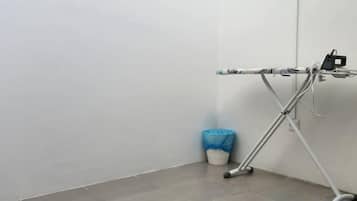 Canto de um quarto com uma cadeira de metal dobrável, um cesto de lixo azul e uma parede branca.