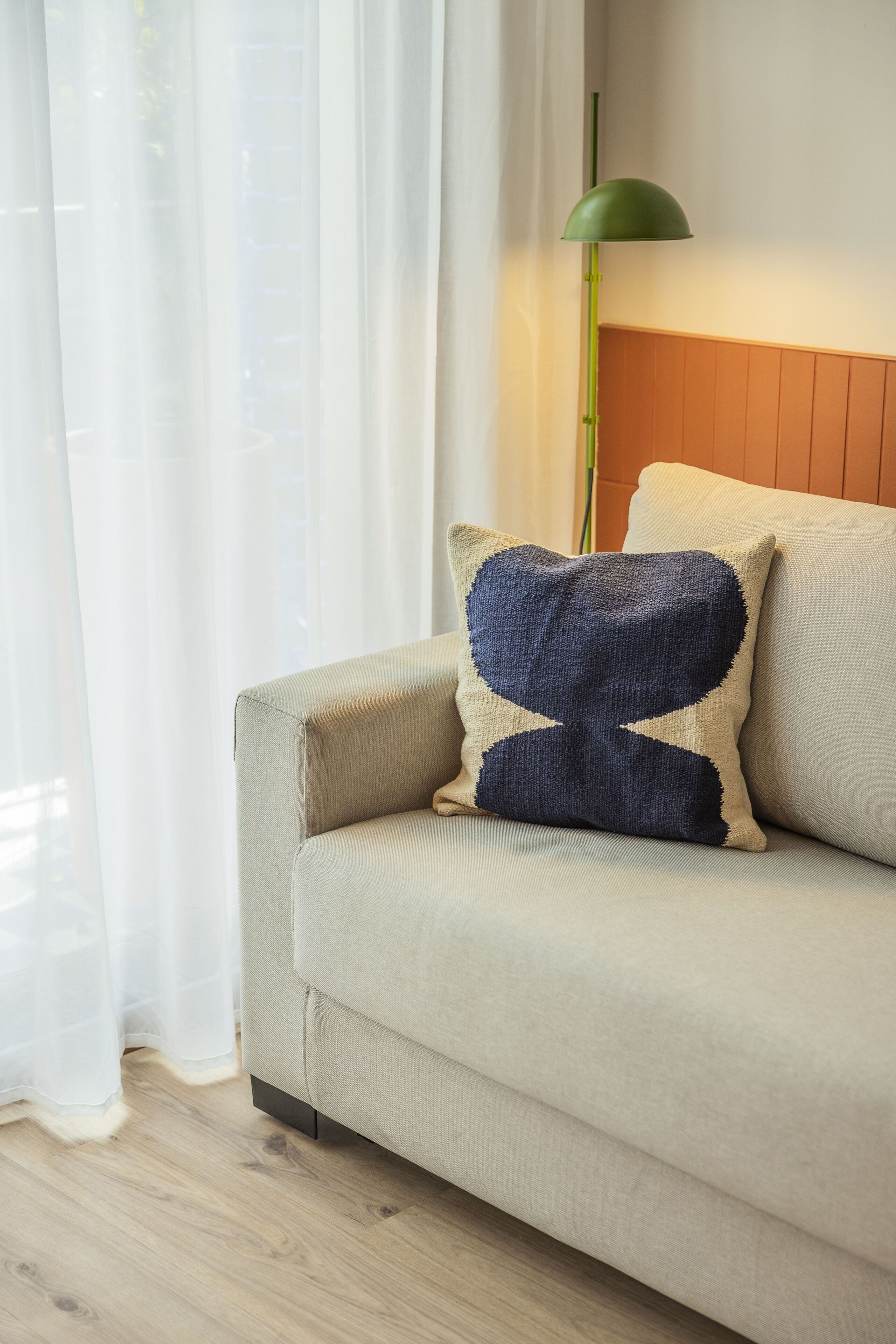 Um sofá bege com um travesseiro azul escuro, um abajur verde e cortinas brancas translúcidas.
