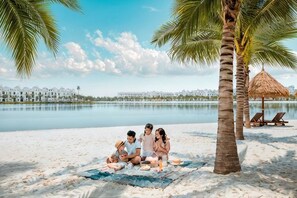 Uma praia com uma família em um cobertor, palmeiras, um guarda-sol de palha e um lago calmo.