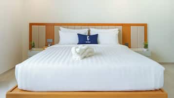 Uma cama bem arrumada com roupa de cama branca e um travesseiro azul, cabeceira de madeira e um pequeno vaso de planta na mesa de cabeceira.