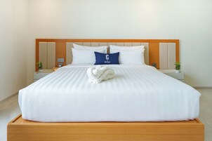 Uma cama bem arrumada com roupa de cama branca e um travesseiro azul, cabeceira de madeira e um pequeno vaso de planta na mesa de cabeceira.