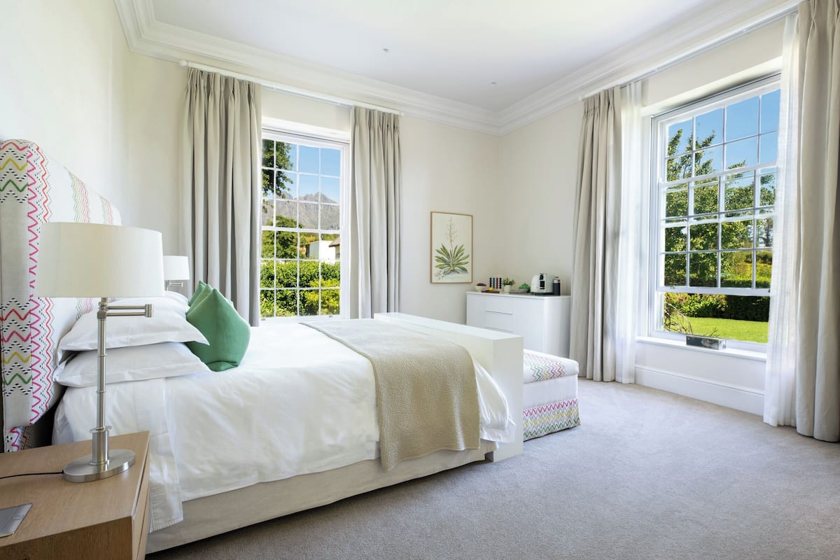 Um quarto espaçoso com uma cama grande, mesa de cabeceira, escrivaninha e vista para o verde pelas janelas.