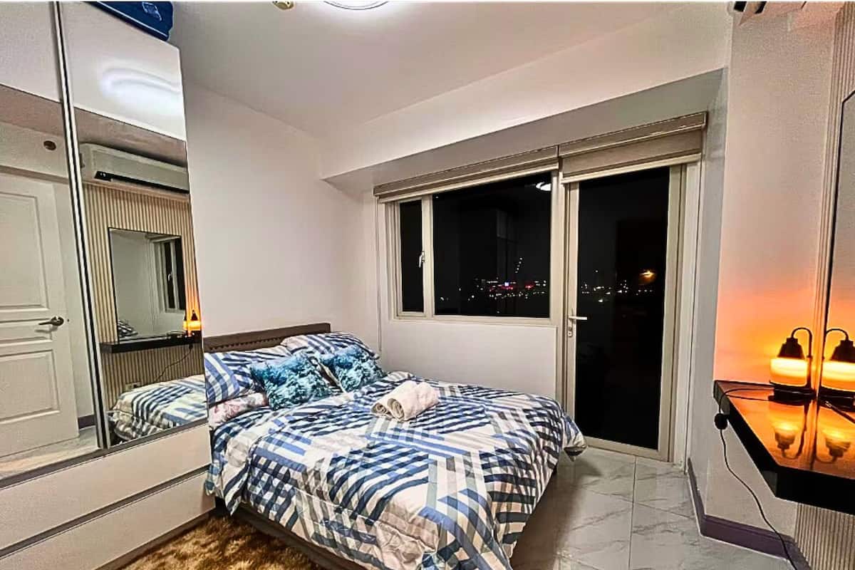 Um quarto com cama, espelho, janela e mesa de cabeceira com abajur.