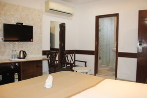 Quarto de hotel com cama, escrivaninha com televisão, espelho, chuveiro e porta.