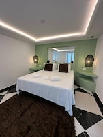 Quarto de hotel moderno com uma cama grande, roupa de cama branca, duas mesas de cabeceira com abajures, um espelho grande e um chão quadriculado em preto e branco.