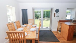 Uma área de refeições com uma mesa de madeira posta para seis pessoas, rodeada por cadeiras de madeira. Uma cozinha com pia e armários é visível à direita.