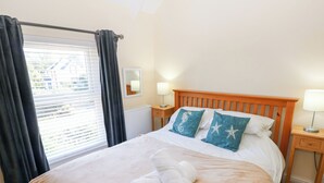 Cama bem arrumada com lençóis brancos e duas almofadas decorativas, cabeceira de madeira, mesas de cabeceira com abajures, janela com cortinas e um radiador.