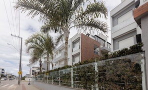 Uma rua urbana moderna com edifícios residenciais, sinal de travessia para pedestres e uma cerca de vidro com vegetação.