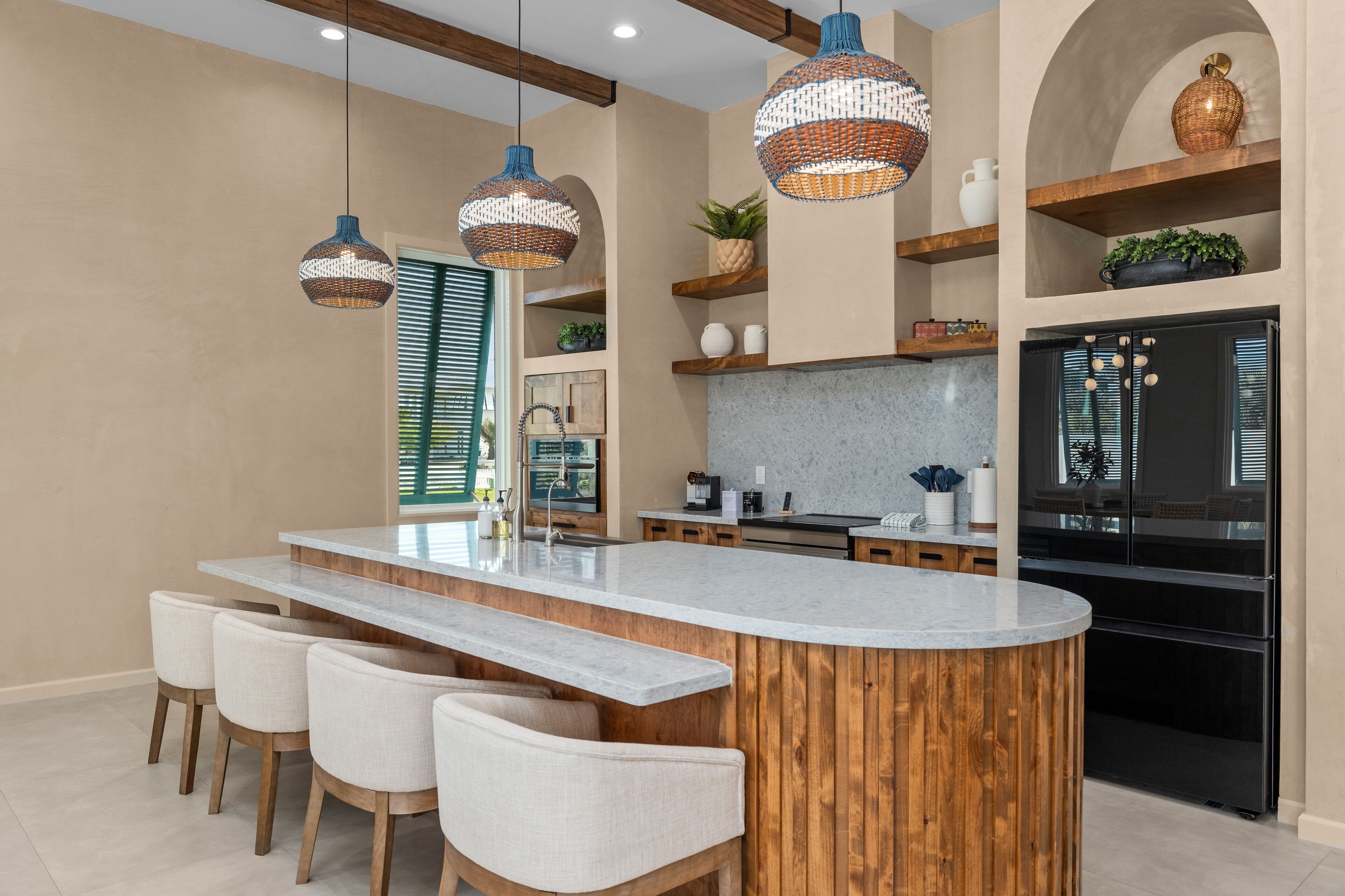 Uma cozinha moderna com ilha central, banquetas, luminárias pendentes de vime e prateleiras embutidas com objetos decorativos.