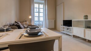 Uma sala de jantar com uma mesa posta para dois, uma TV em um armário branco e uma janela com cortinas.