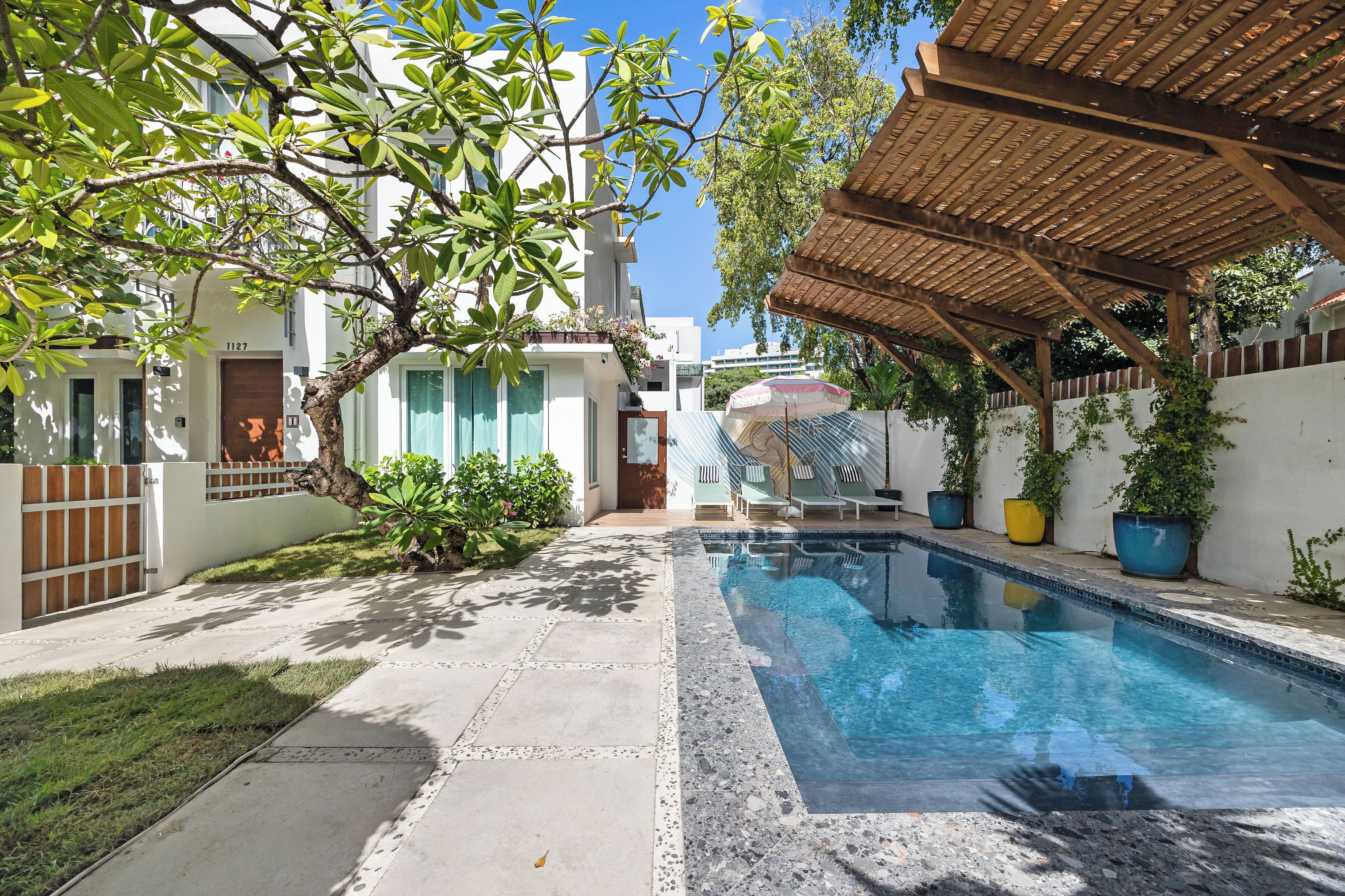 Piscina com pérgola de madeira, rodeada por um edifício branco com relvado verde e plantas em vasos.