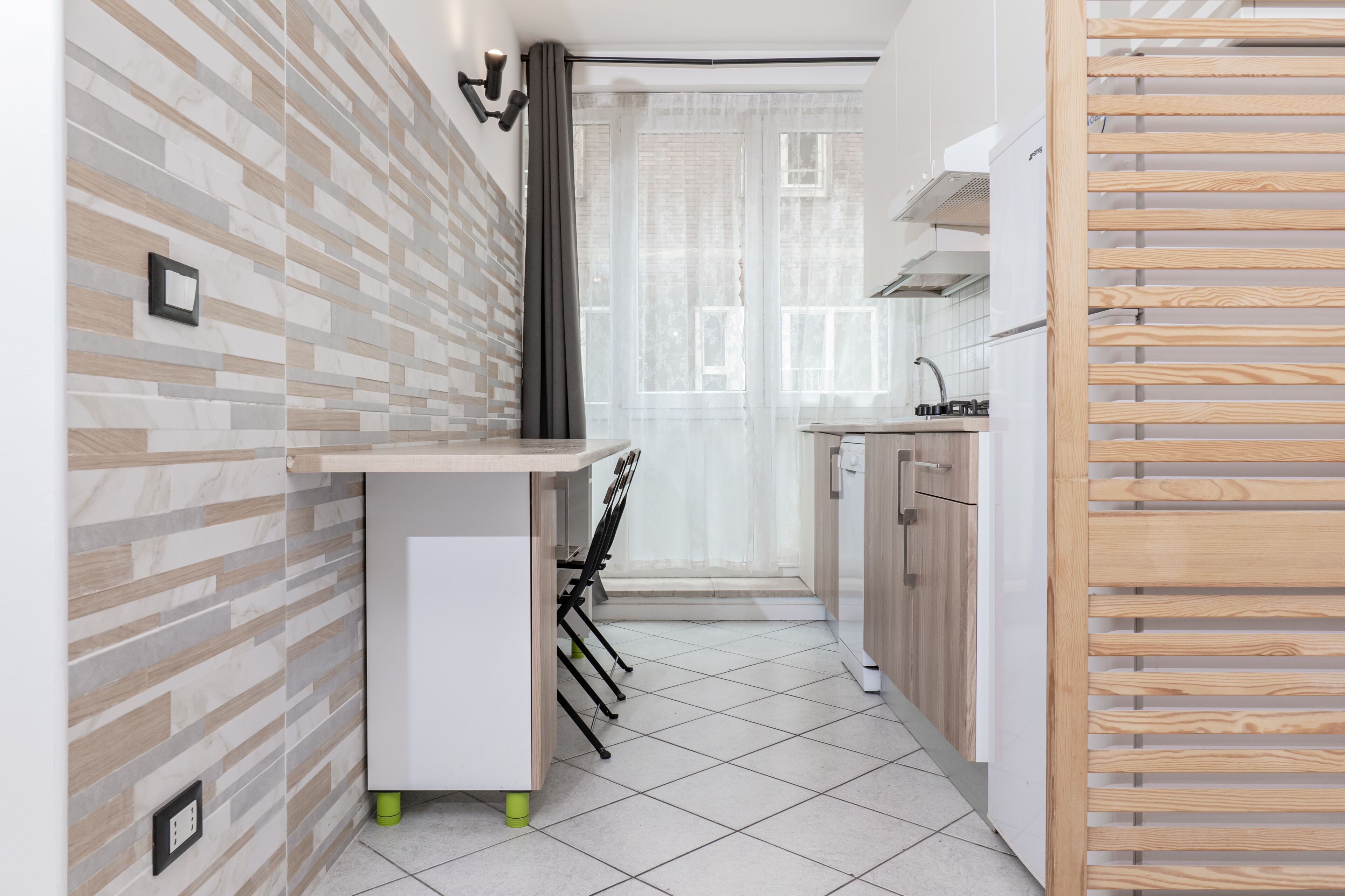 Uma cozinha compacta com chão em azulejo, bancada embutida e janela com cortinas leves.