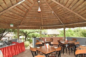 Uma área de refeições ao ar livre com mesas e cadeiras de madeira, um serviço de buffet e uma estrutura com telhado de palha ao fundo.