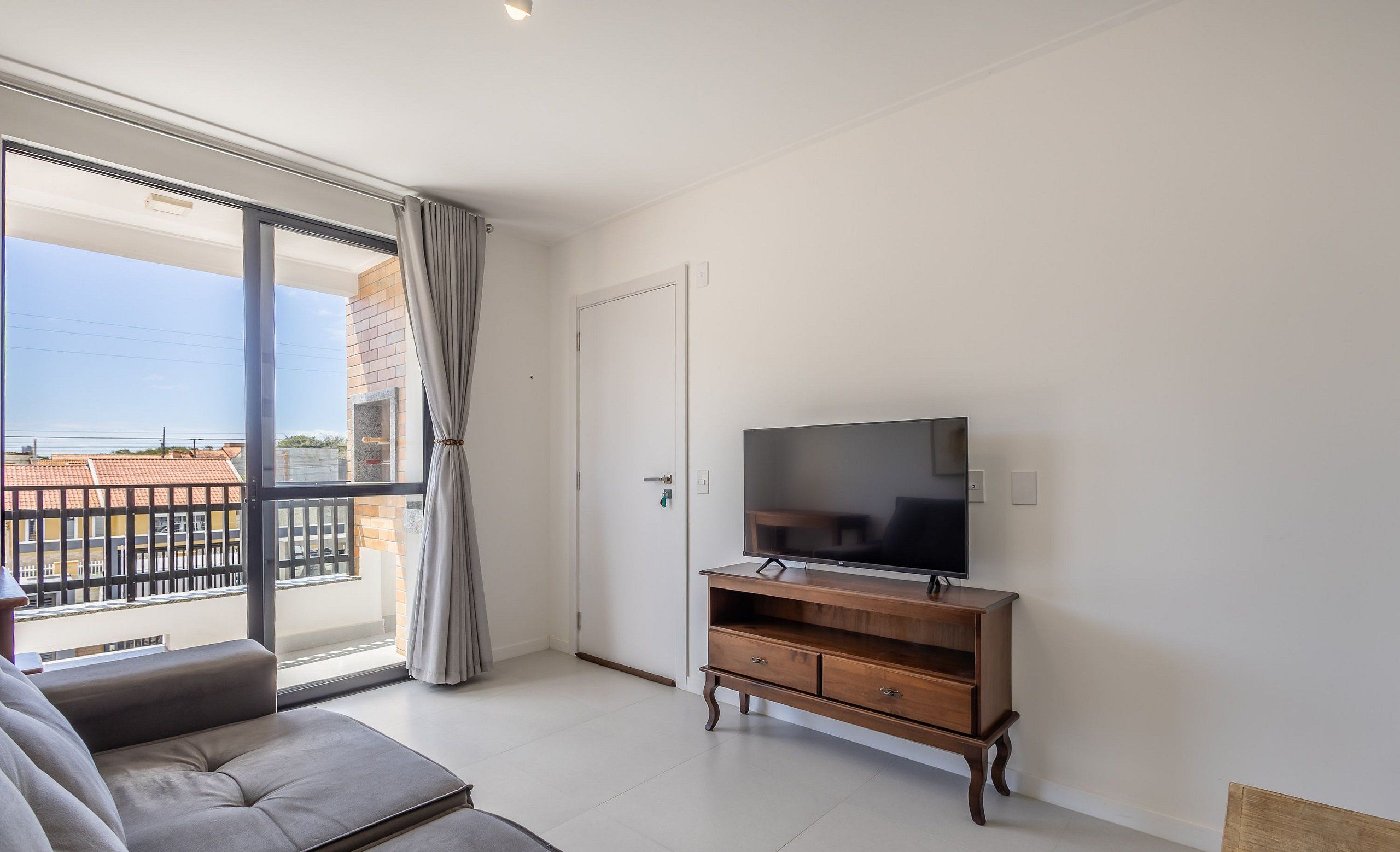 Uma sala de estar moderna com sofá, televisão sobre um suporte de madeira e porta de vidro deslizante para uma varanda.