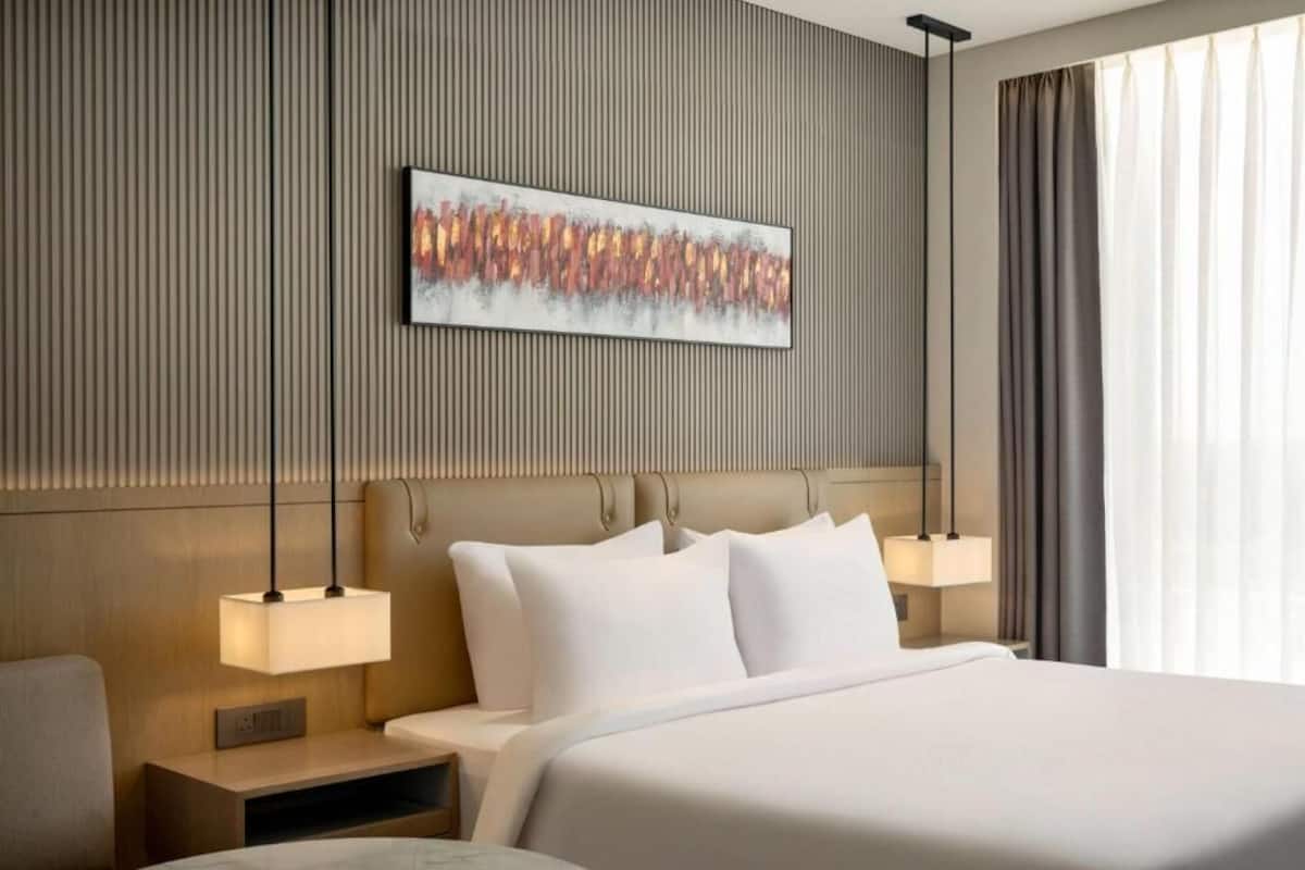 Quarto de hotel com cama, abajures, cabeceira, obra de arte na parede e janela com cortinas.