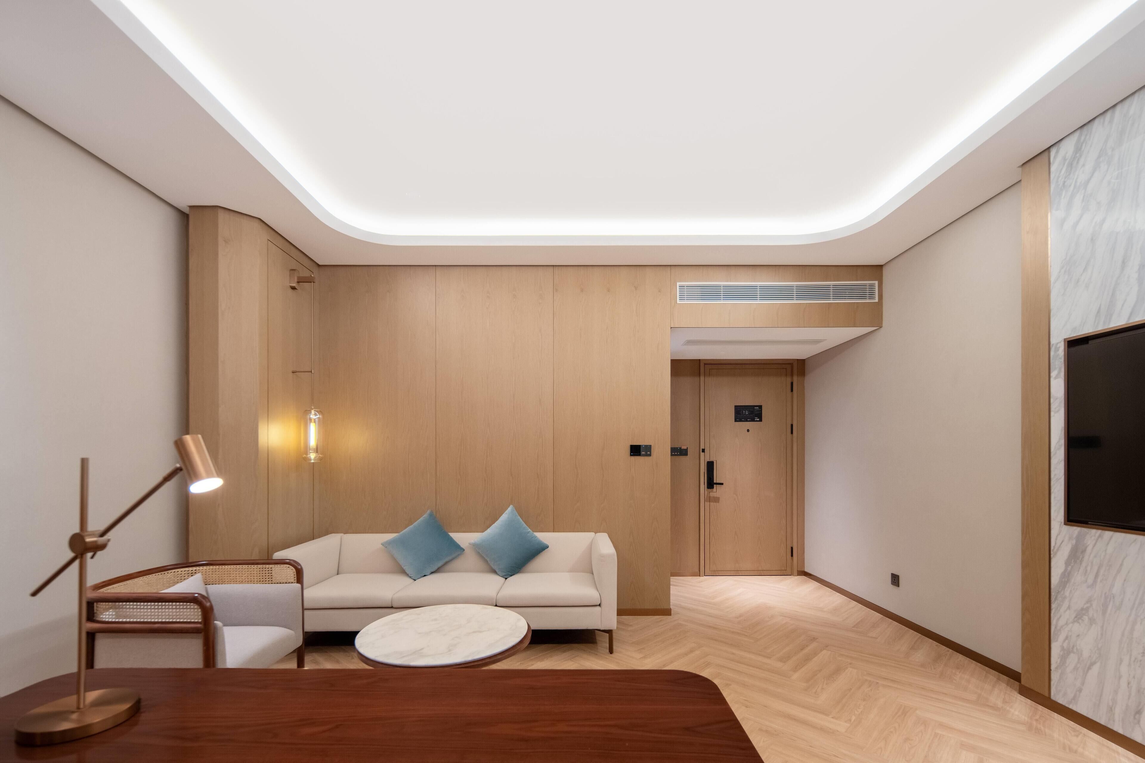 Quarto de hotel moderno com parede de painel de madeira, sofá branco com almofadas azuis, mesa de centro redonda e TV fixada na parede.