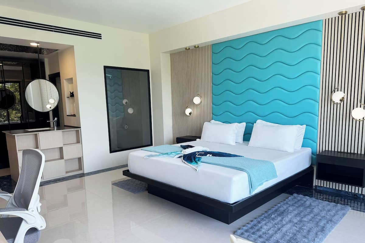 Quarto de hotel moderno com uma cama grande, uma escrivaninha com cadeira, um banheiro com chuveiro de vidro e vista para o exterior.