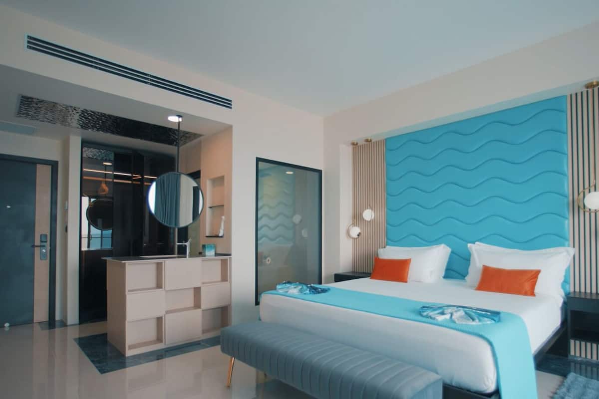 Quarto de hotel moderno com uma cama, uma toucador com espelho, um guarda-roupa e um banheiro visível através de uma porta de vidro.