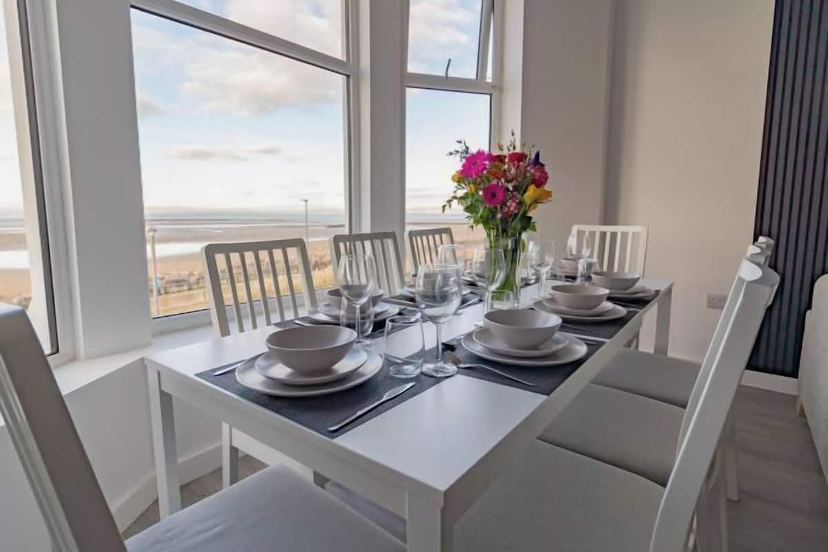 Mesa de jantar posta para seis com cadeiras brancas, um vaso de flores e vista para a praia.
