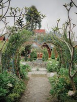 Um caminho no jardim leva a um edifício com telhado vermelho, rodeado por vegetação exuberante e um arco de metal decorativo.