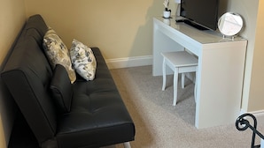 Quarto com um sofá preto, uma escrivaninha branca e uma televisão.