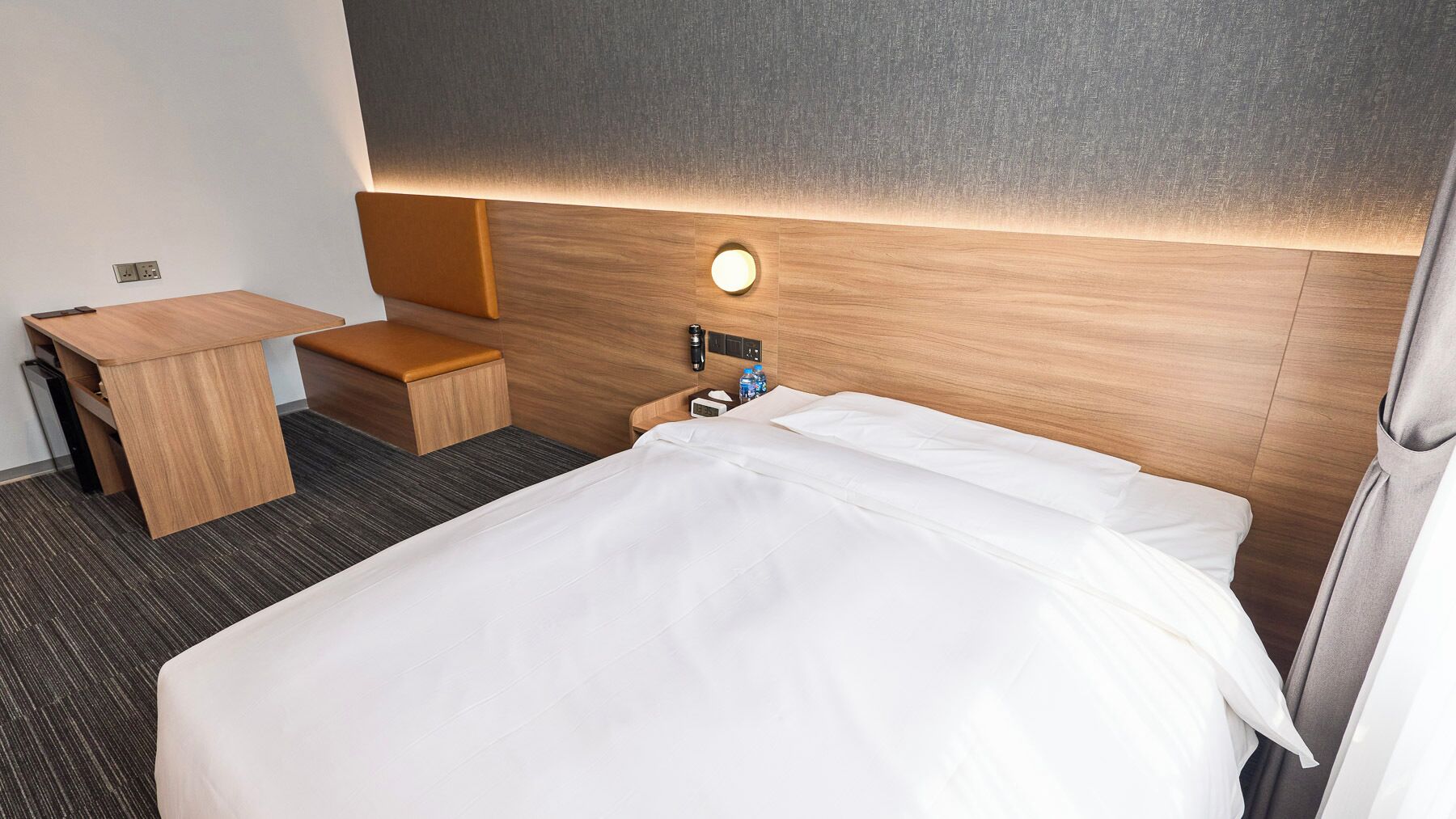 Quarto de hotel com cama, escrivaninha, cadeira, mesa de cabeceira e luminária na parede.