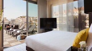 Quarto de hotel com uma cama grande, televisão e varanda com cadeiras de descanso e vista para a cidade.